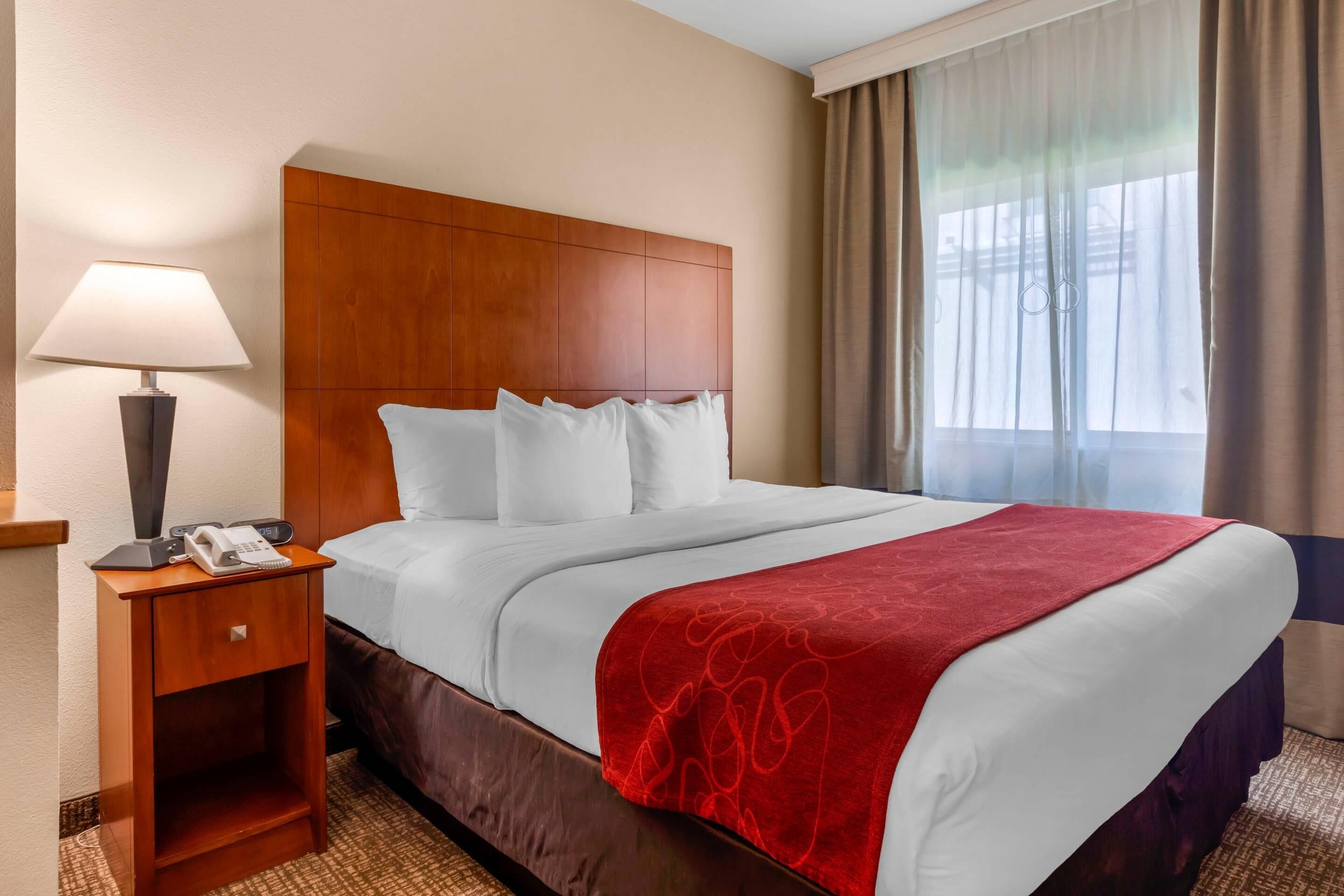 Quarto de hotel com cabeceira de madeira, cama branca com manta vermelha, mesa de cabeceira com abajur e telefone, e janela com cortinas.