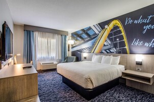 Quarto de hotel com cama, mesas de cabeceira, escrivaninha, cadeira, televisão e um grande painel decorativo na parede.