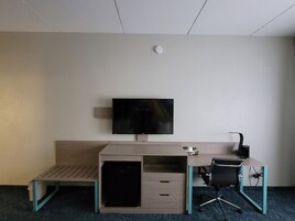 Quarto de hotel com escrivaninha, uma cadeira, televisão fixada na parede e um pequeno cesto de lixo.