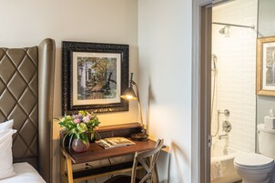 Quarto de hotel com cama, escrivaninha com luminária, quadro emoldurado e banheiro com chuveiro e vaso sanitário.