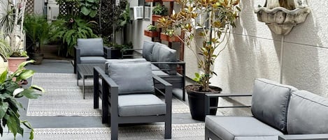 Um pátio externo com sofás e cadeiras cinzas, um tapete estampado e plantas em vasos.
