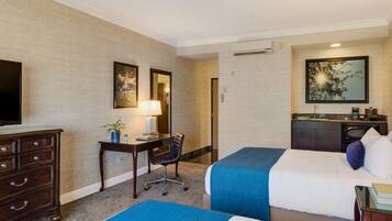 Quarto de hotel com duas camas, uma secretária com cadeira, televisão e uma kitchenette pequena.