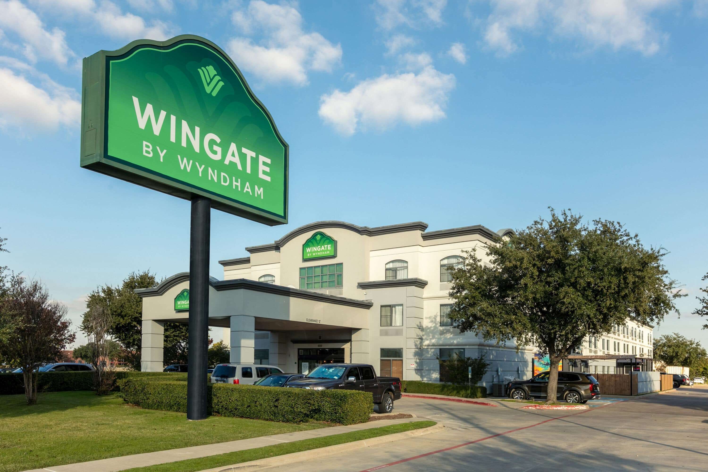 Um hotel Wingate by Wyndham com uma grande placa, área de estacionamento e um edifício com várias janelas e entrada coberta.