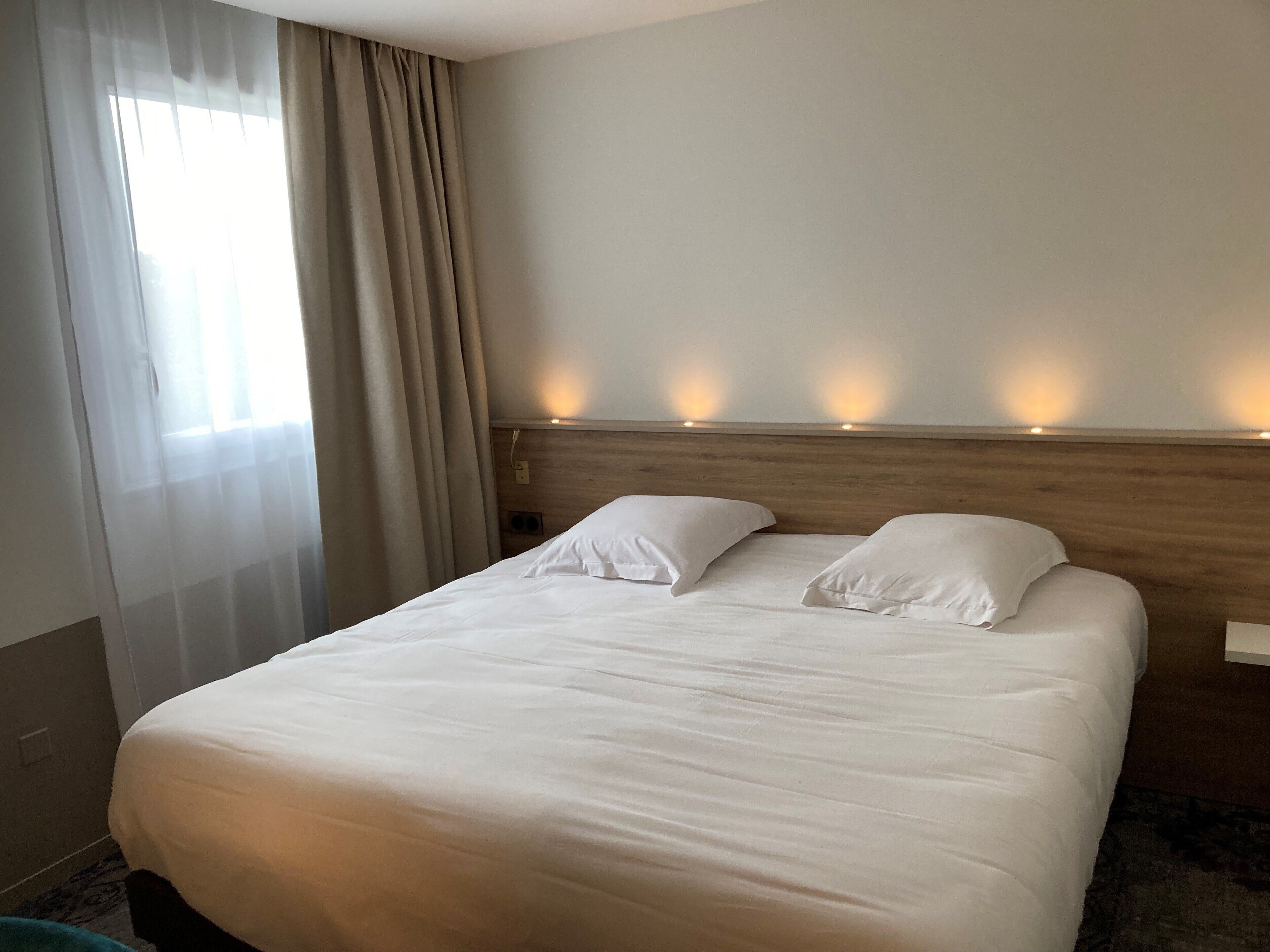 Quarto de hotel com cama grande, cortinas bege e luminárias fixadas na parede.
