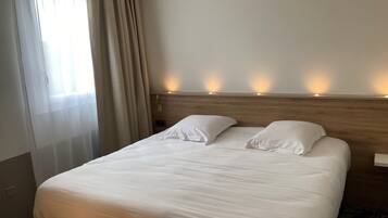 Quarto de hotel com cama grande, cortinas bege e luminárias fixadas na parede.