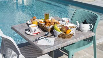 Disposição de café da manhã junto à piscina, com croissants, frutas e bebidas.