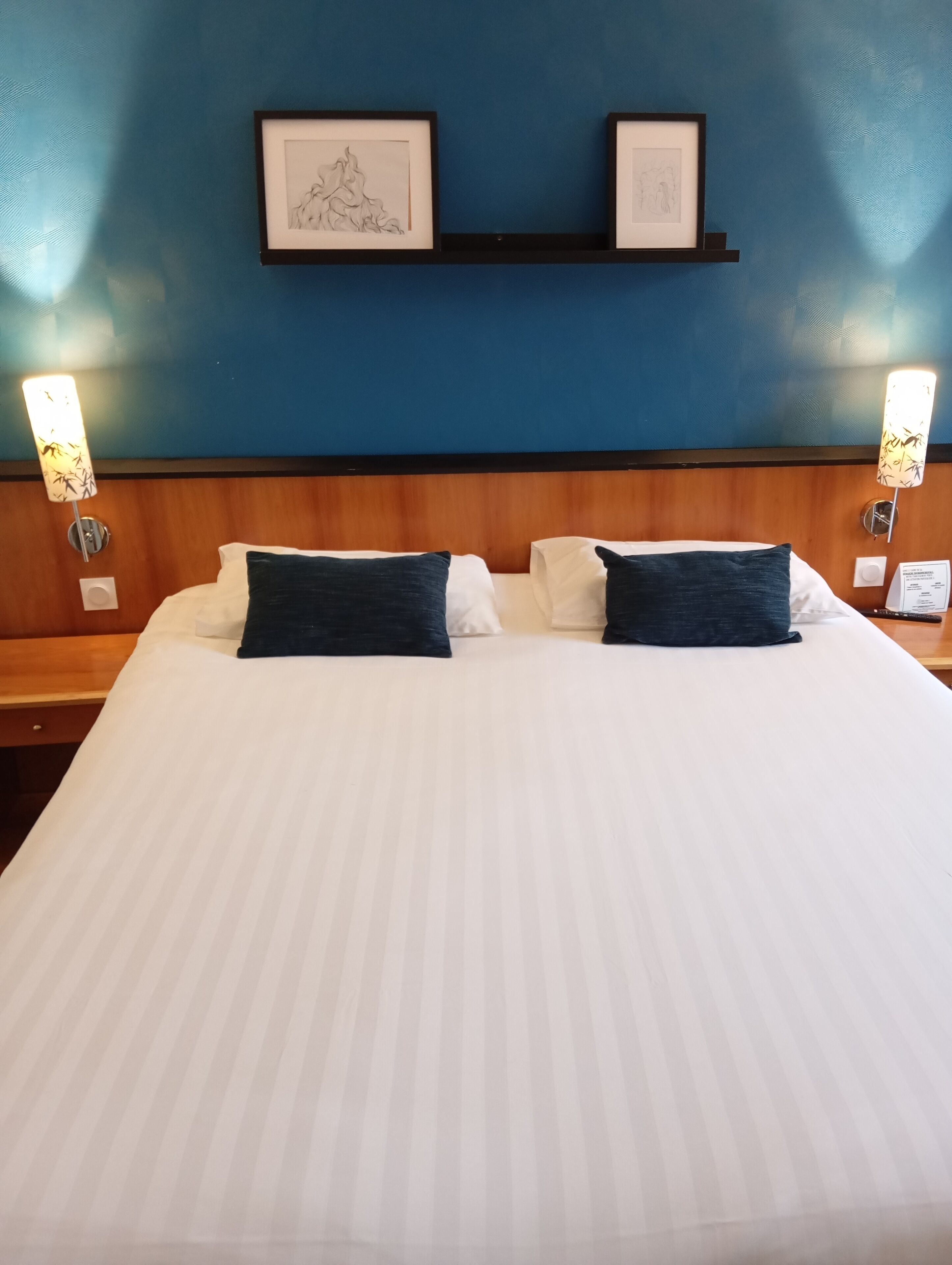 Quarto de hotel com uma cama grande, duas luminárias fixadas na parede e duas obras de arte emolduradas.