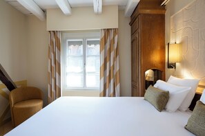 Quarto de hotel com uma cama grande, uma cadeira, uma janela com cortinas e uma luminária na parede.