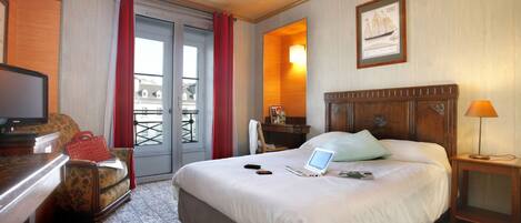 Quarto de hotel com cama, escrivaninha com laptop, cadeira, televisão e janela com cortinas.