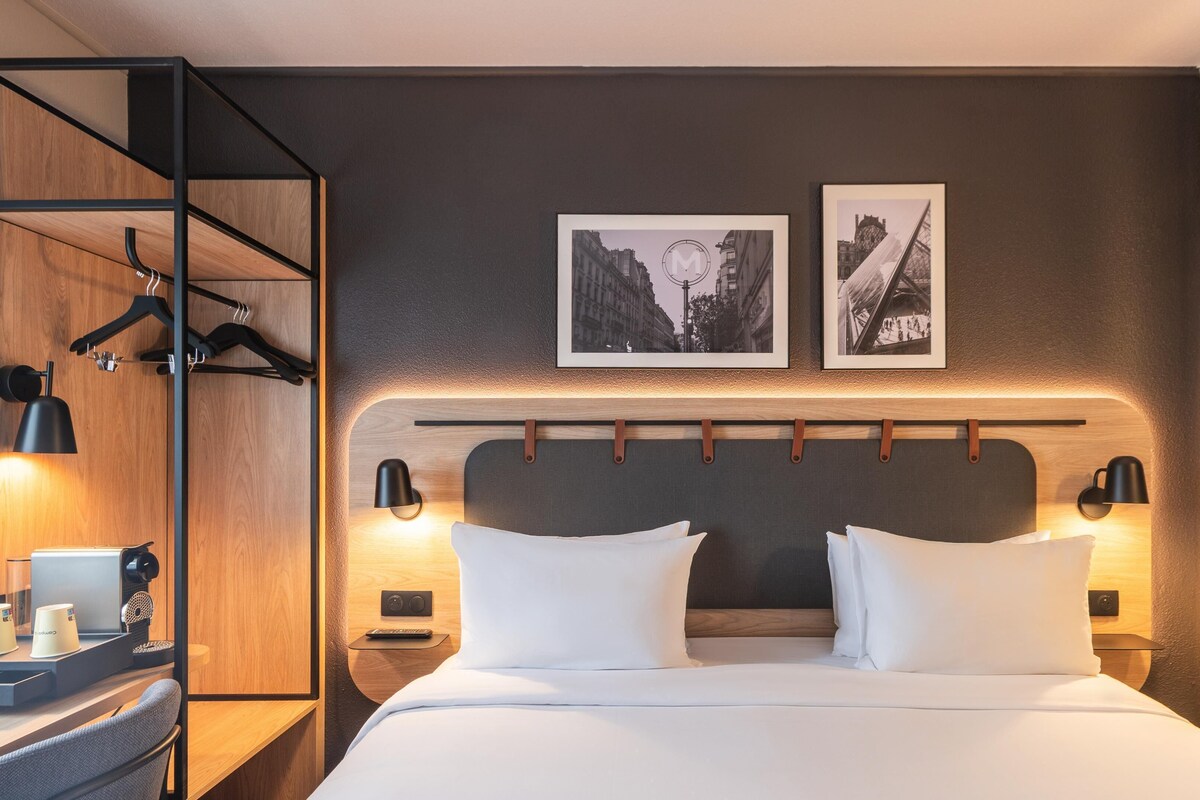 Quarto de hotel com cama, abajures, guarda-roupa de madeira, escrivaninha com luminária e duas quadros pendurados na parede.
