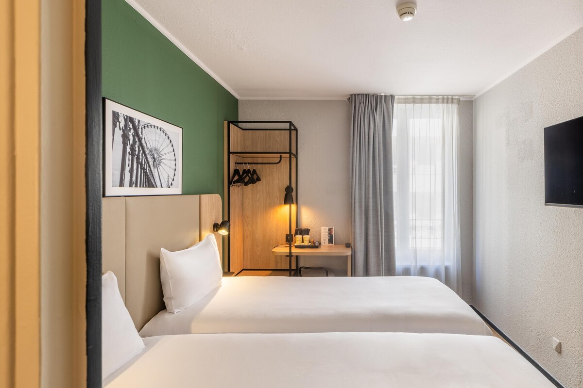Quarto de hotel com duas camas, uma escrivaninha com abajur, um cabideiro, um quadro emoldurado e uma televisão.
