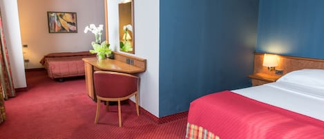 Quarto de hotel com cama, uma escrivaninha com cadeira, uma mesinha com um vaso de flores, espelho e um quadro na parede.