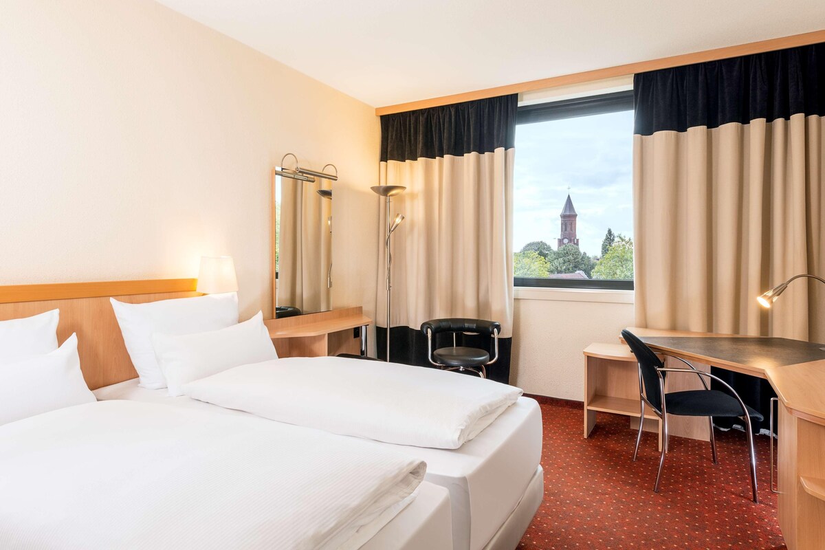 Quarto de hotel com duas camas, uma mesa, uma cadeira, chuveiro e janela com vista para uma torre.