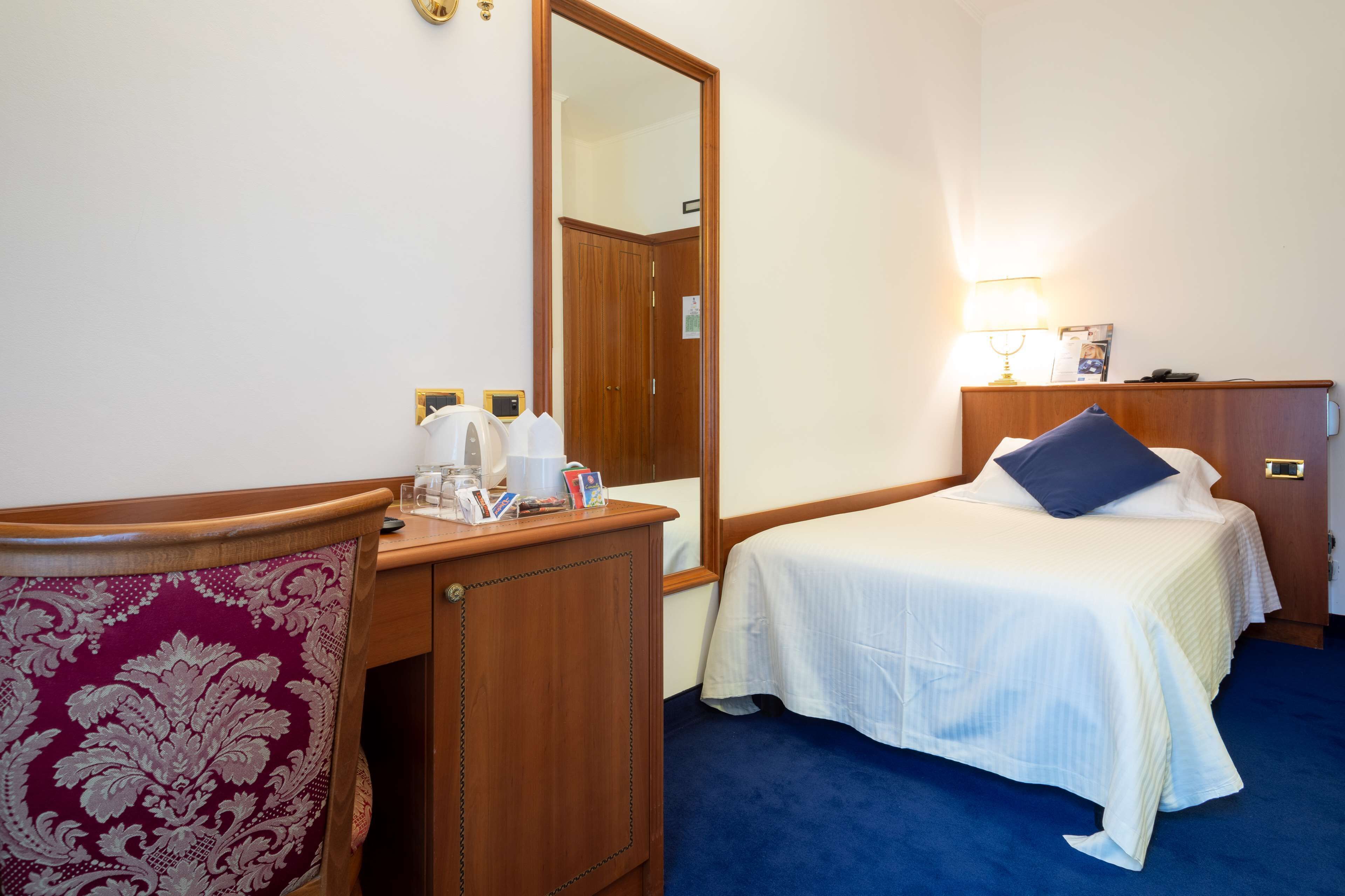 Quarto de hotel com cama, mesa de cabeceira de madeira, espelho e escrivaninha com cadeira.