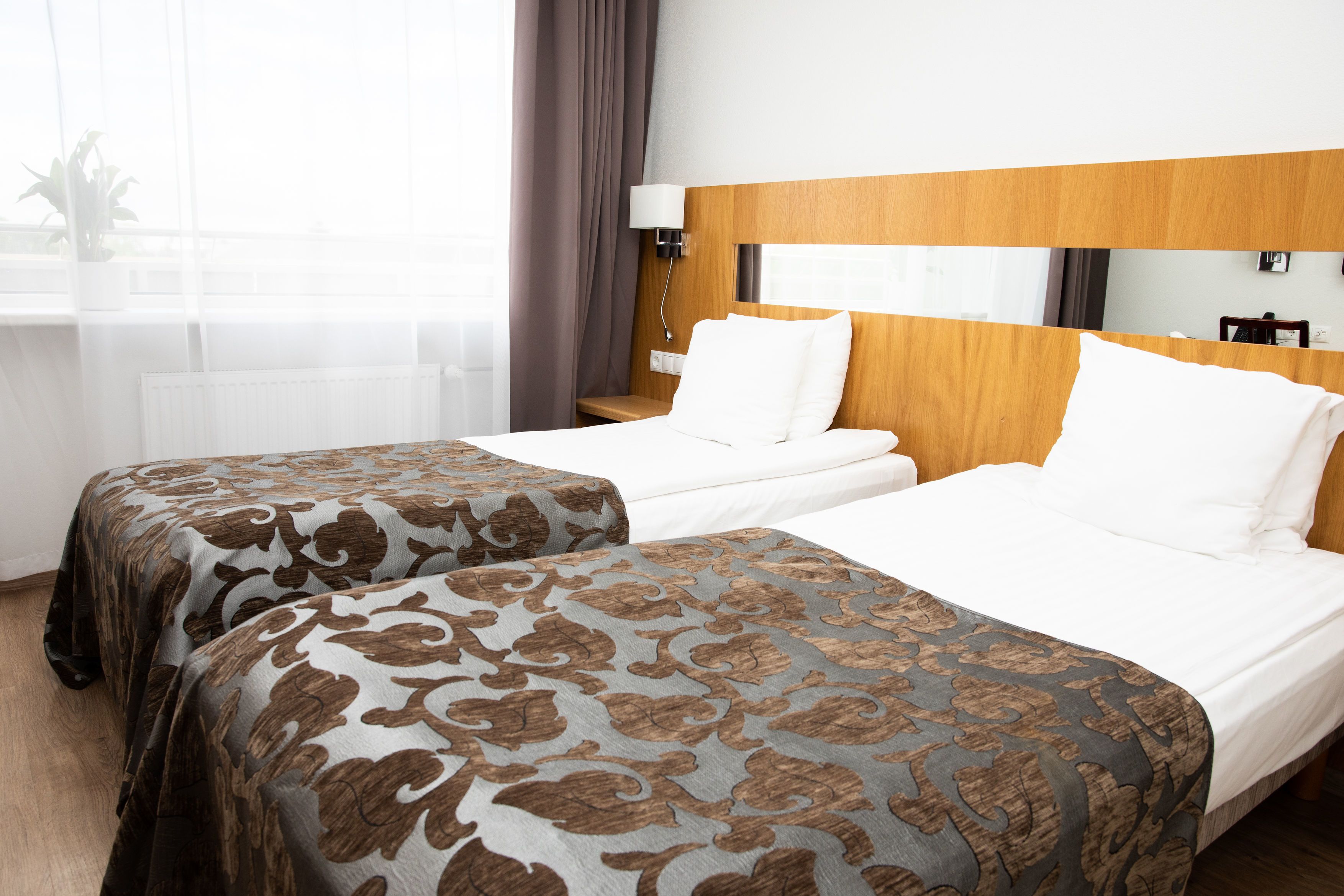 Quarto de hotel com duas camas, cabeceira de madeira, mesa de cabeceira com abajur e janela com cortinas.