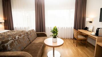 Quarto de hotel com cama, mesa redonda com planta, escrivaninha com cadeira e televisão na parede.