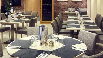 Um restaurante com mesas redondas, cadeiras cinza estofadas e uma parede de tijolos com quadros emoldurados.