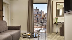 Quarto de hotel com sofá, duas mesinhas redondas com bebidas, vista da cidade pela janela com cortinas e uma pequena mesa com flores.