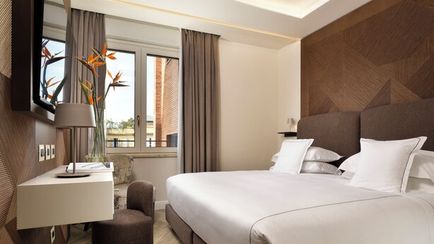 Quarto de hotel moderno com uma cama grande, uma escrivaninha com luminária e vista para a cidade pela janela.
