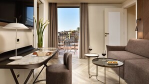 Quarto de hotel com área de refeições, um sofá, uma mesa com tampo de vidro, uma luminária e vista para a cidade.