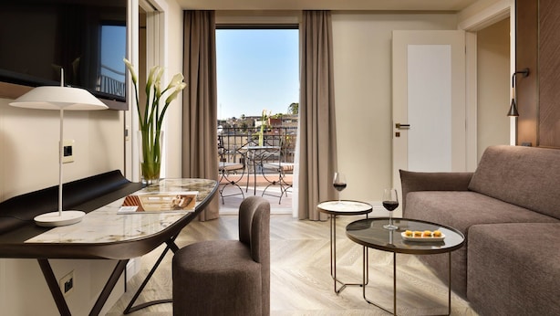 Quarto de hotel com área de refeições, um sofá, uma mesa com tampo de vidro, uma luminária e vista para a cidade.