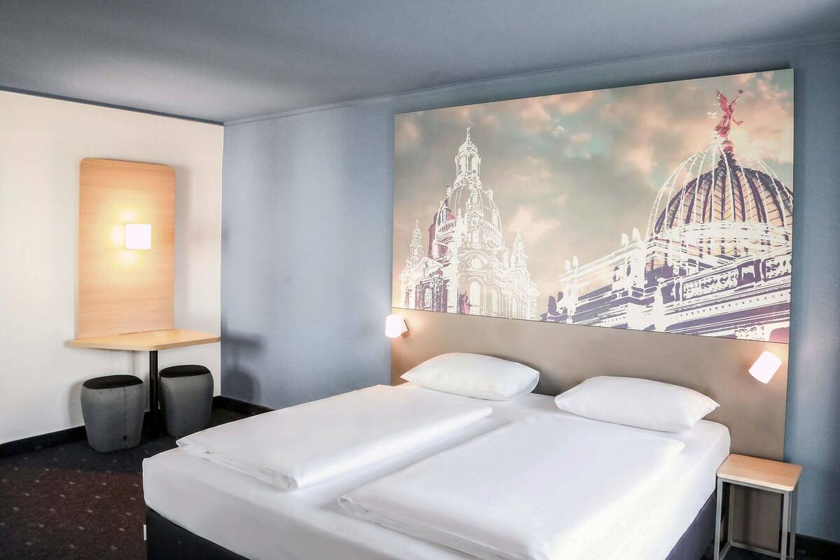 Quarto de hotel com uma cama, duas mesas de cabeceira, uma luminária na parede e uma grande obra de arte na parede retratando um edifício.