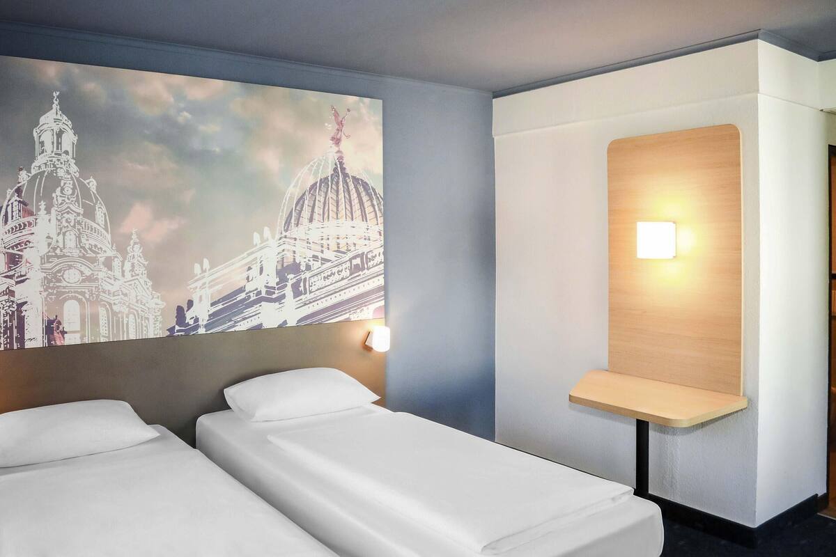 Quarto de hotel com duas camas, uma luminária na parede e um mural de um edifício.