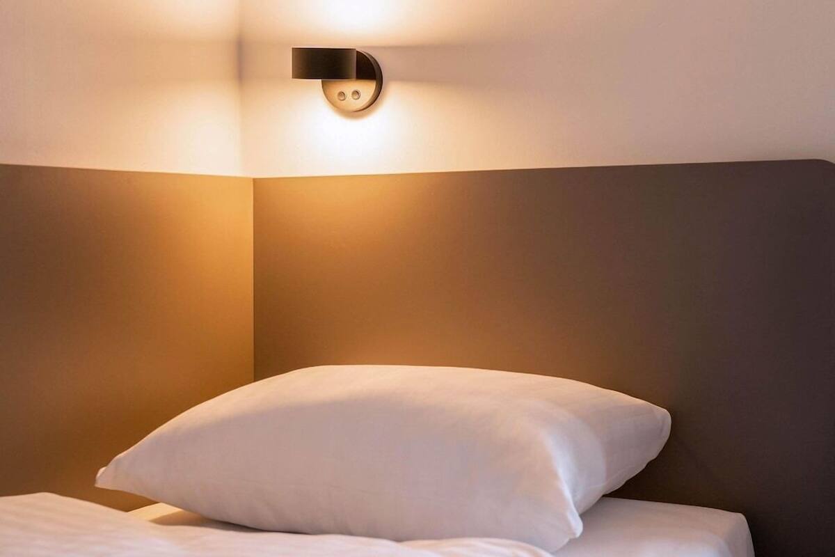 Quarto de hotel com cama, travesseiro e luminária fixada na parede.
