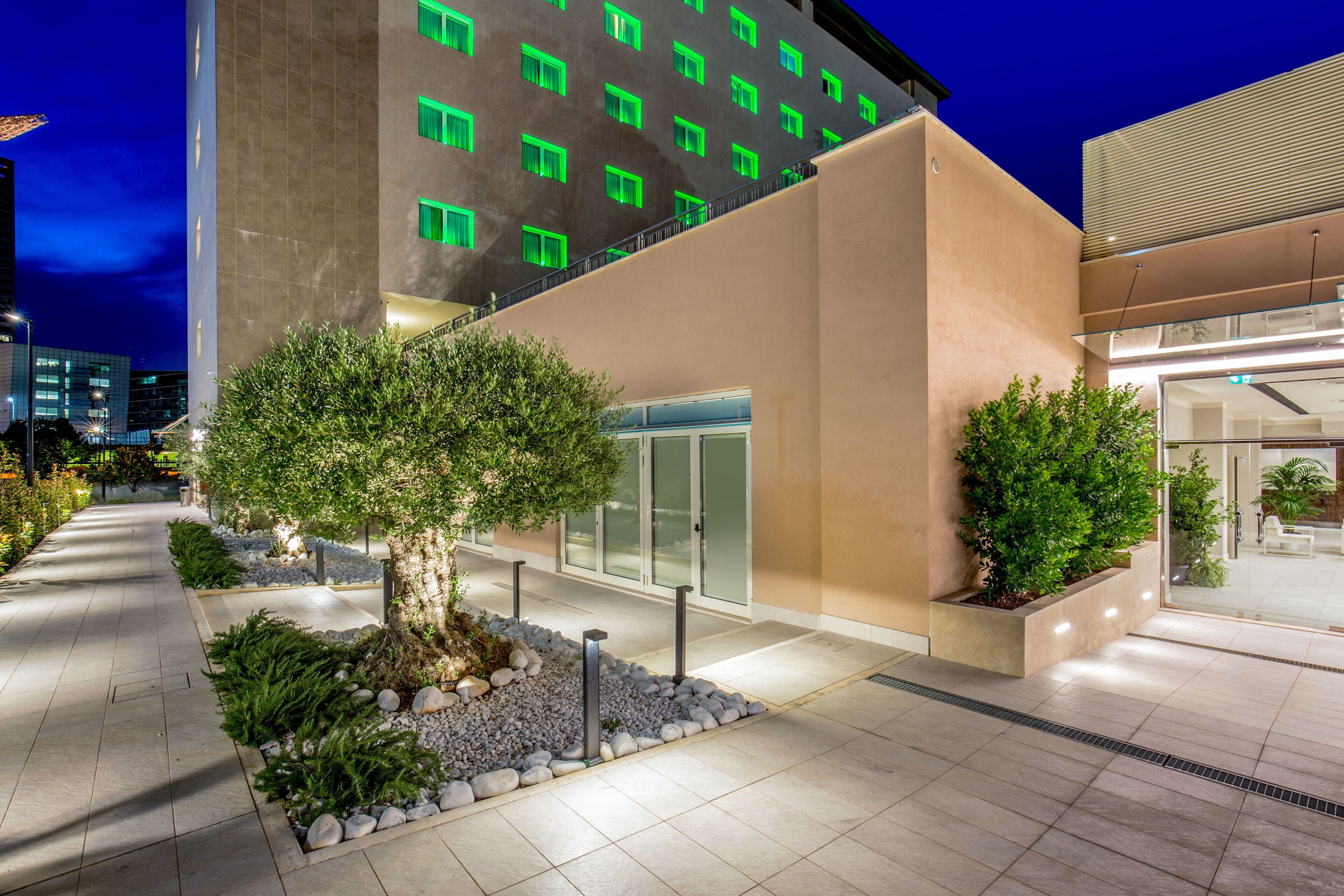 Exterior de um hotel moderno com um jardim bem cuidado, um caminho e um edifício com várias janelas.