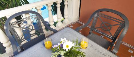 Uma mesa com duas cadeiras, um vaso de flores e dois copos de sumo de laranja num terraço com piscina ao fundo.
