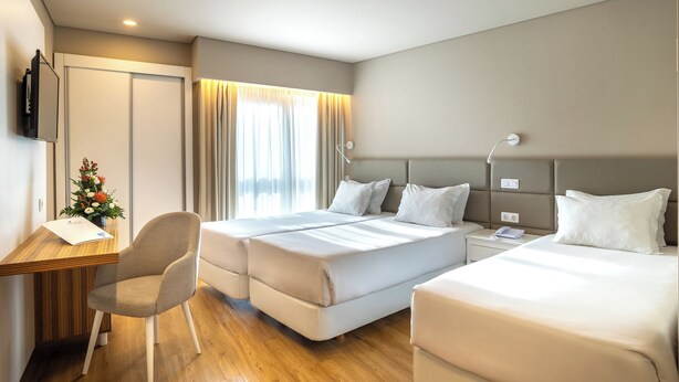 Quarto de hotel com duas camas, uma escrivaninha, uma cadeira, uma televisão e uma janela com cortinas.
