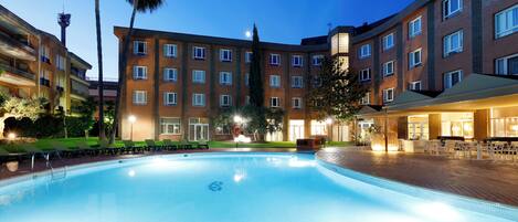 Área de piscina de hotel com céu claro, palmeiras e um edifício com várias janelas e varandas.