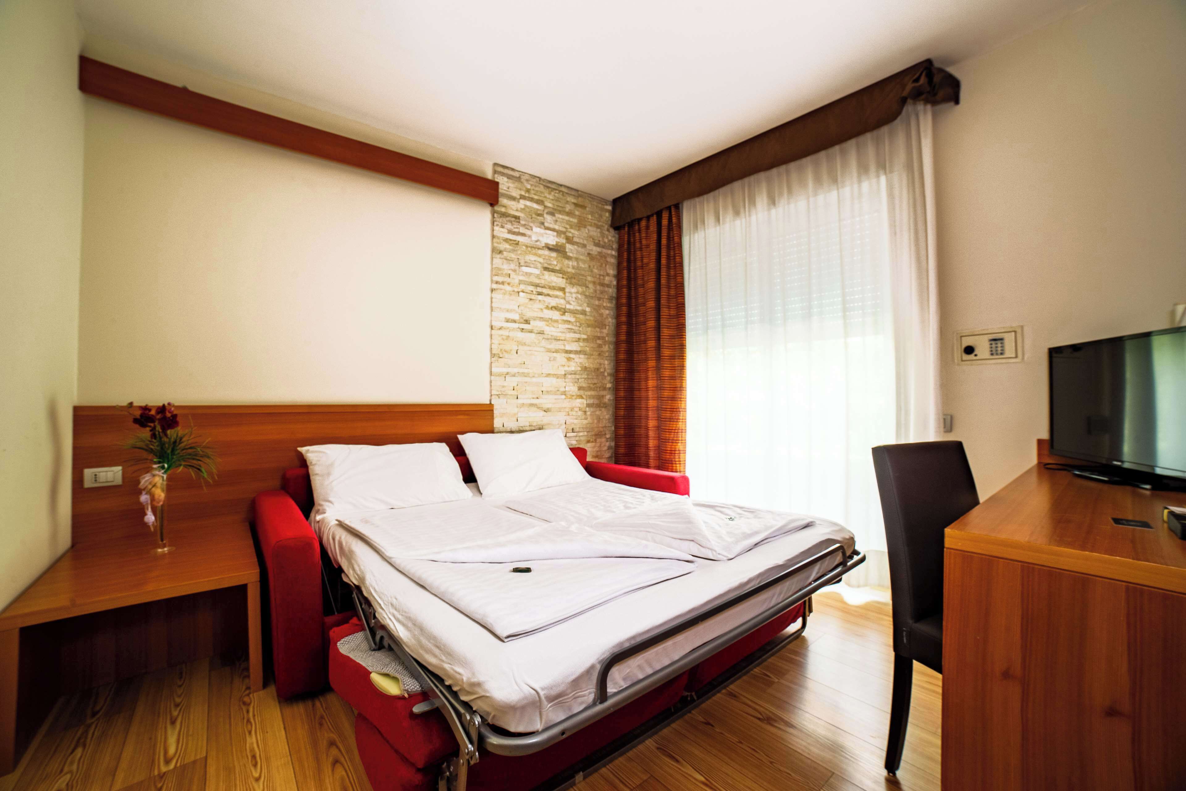 Quarto de hotel com cama, secretária, televisão, cadeira e uma pequena mesa com um vaso de flores.