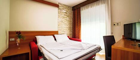 Quarto de hotel com cama, secretária, televisão, cadeira e uma pequena mesa com um vaso de flores.