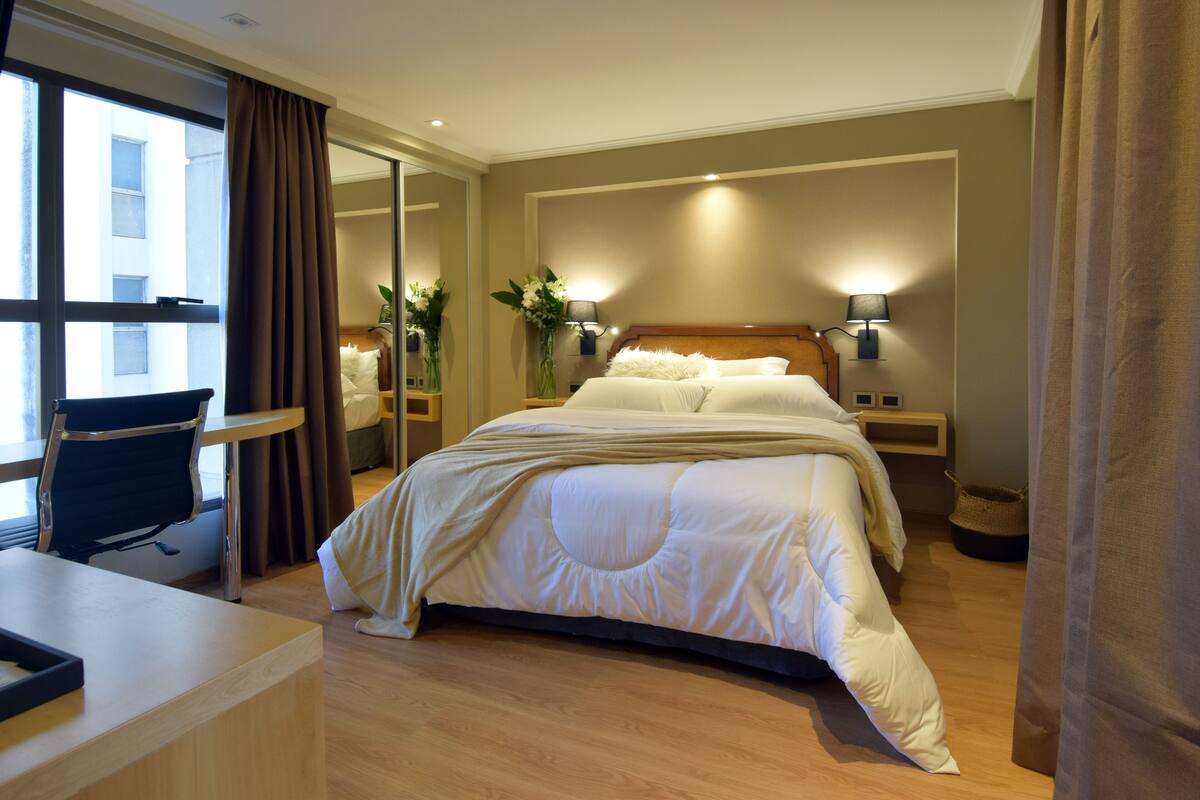 Quarto de hotel com uma cama grande, uma escrivaninha, uma cadeira e uma janela com cortinas.