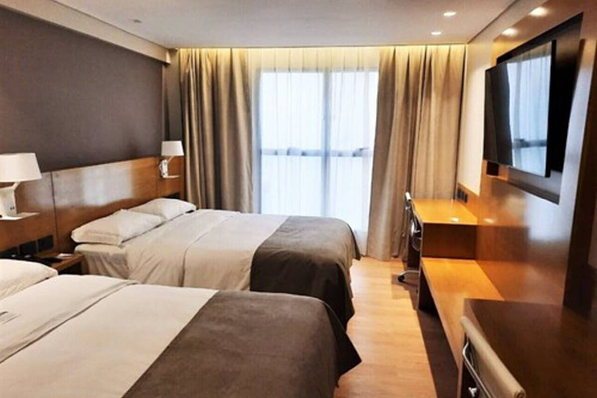 Quarto de hotel com duas camas, uma escrivaninha, televisão e uma janela grande com cortinas.