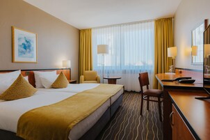 Quarto de hotel com uma cama grande, duas cadeiras, uma mesa de trabalho e uma janela com cortinas.