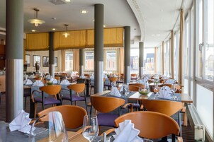 Um restaurante com várias mesas preparadas para refeições, grandes janelas e vista para a cidade.