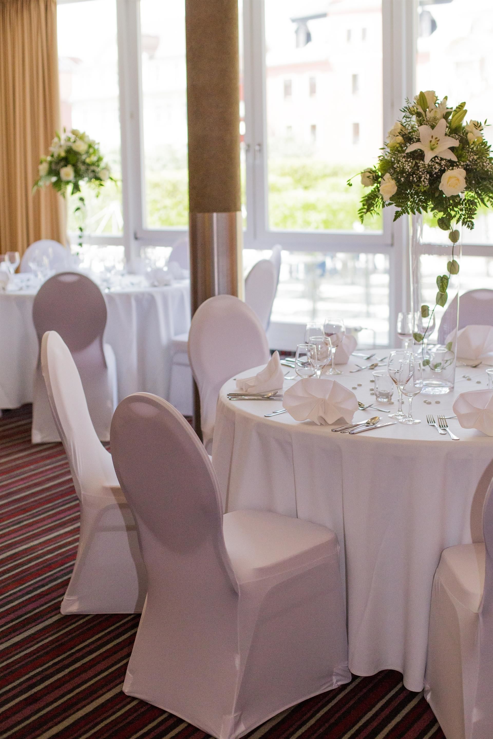 Uma área de jantar com mesas redondas dispostas para um evento formal, cadeiras brancas e centros de mesa florais.