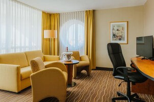 Quarto de hotel com sofá, poltronas, uma pequena mesa com garrafa e copos, televisão e um quadro na parede.