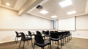 Uma sala de conferências com projetor, fileiras de cadeiras, piso coberto de carpete e parede com quadro branco.