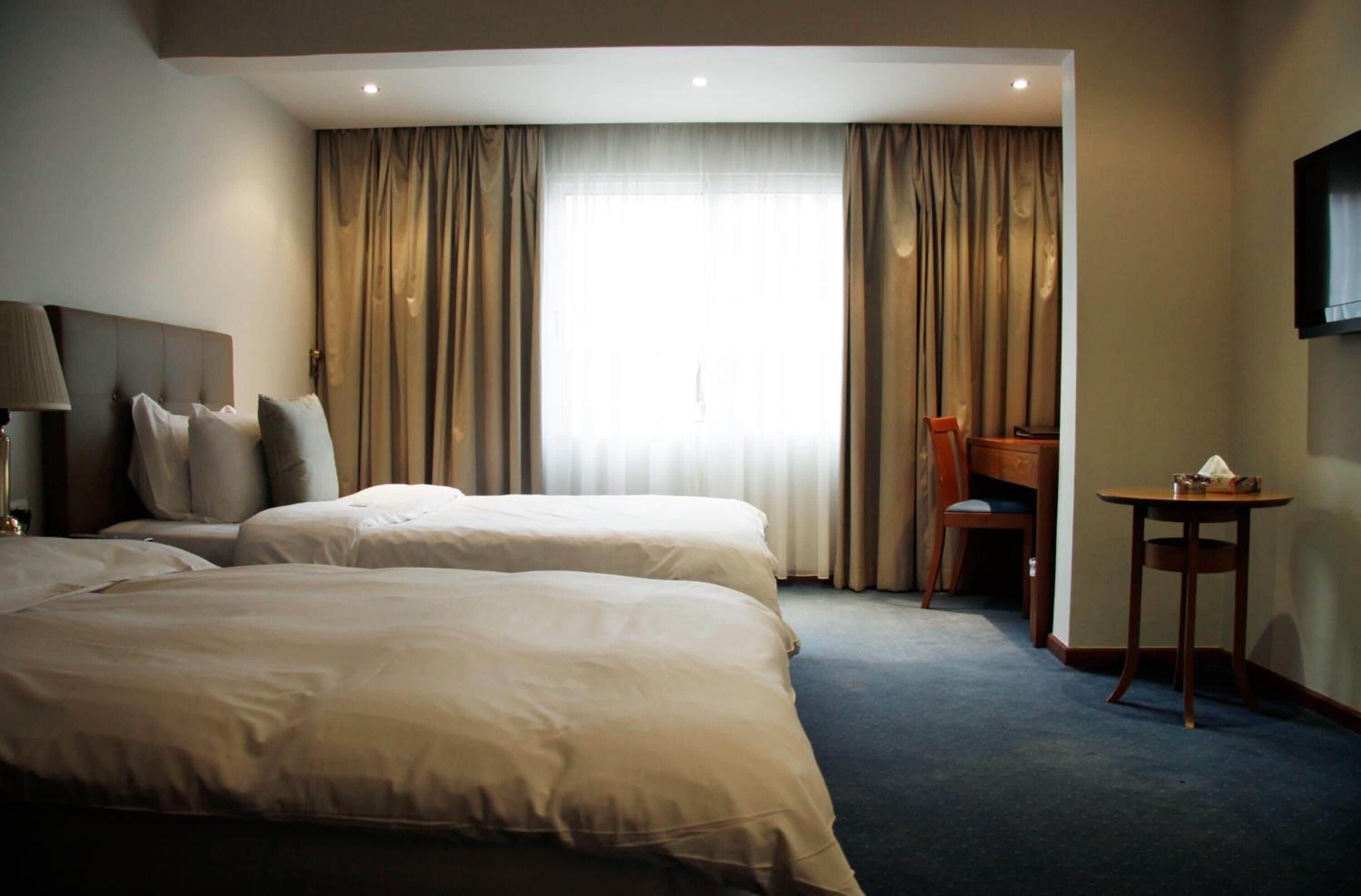 Quarto de hotel com duas camas, uma escrivaninha, uma cadeira, uma televisão e uma janela com cortinas.