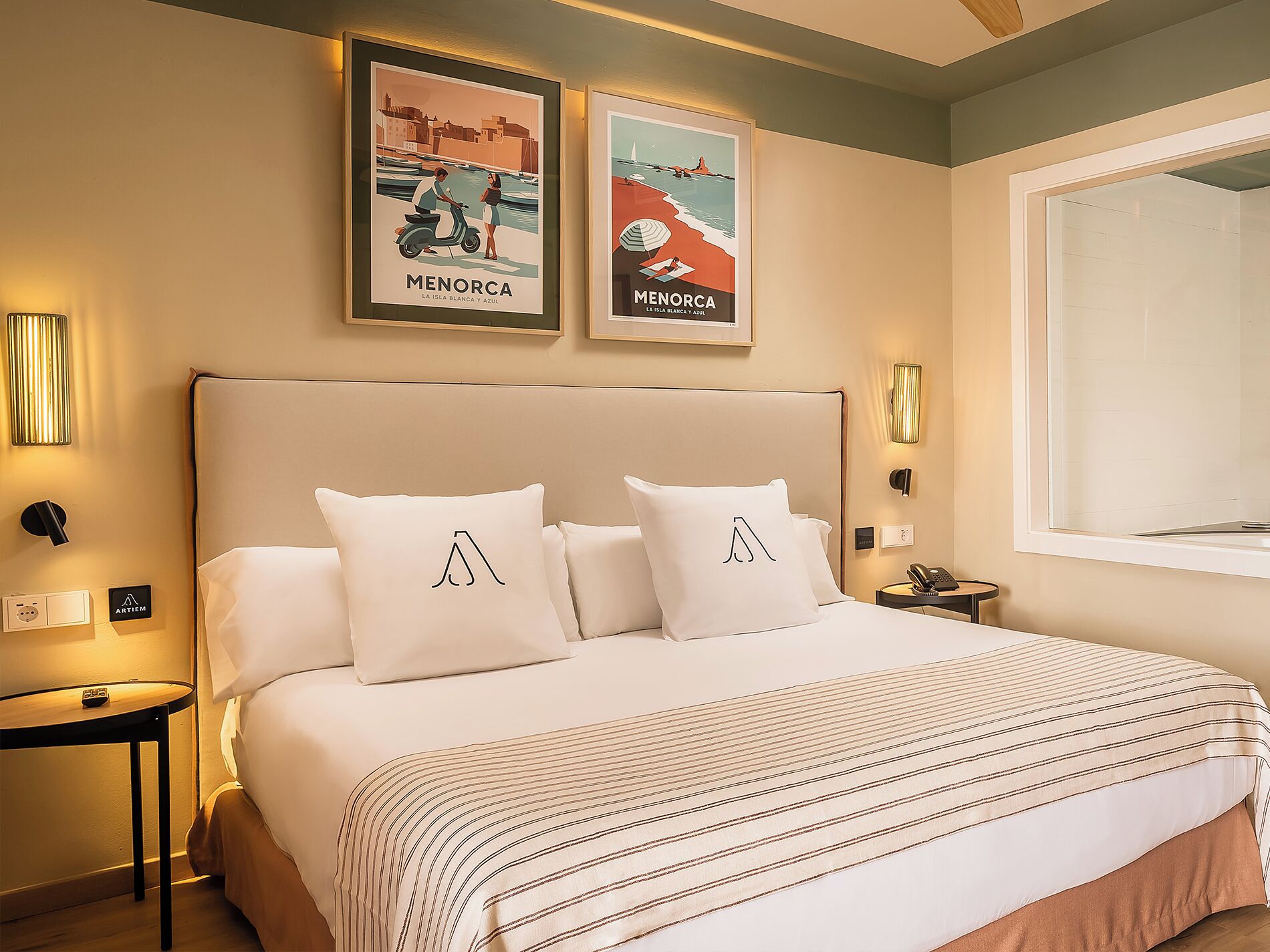 Quarto de hotel com uma cama grande, dois quadros na parede, mesas de cabeceira com abajures e uma janela com vista.