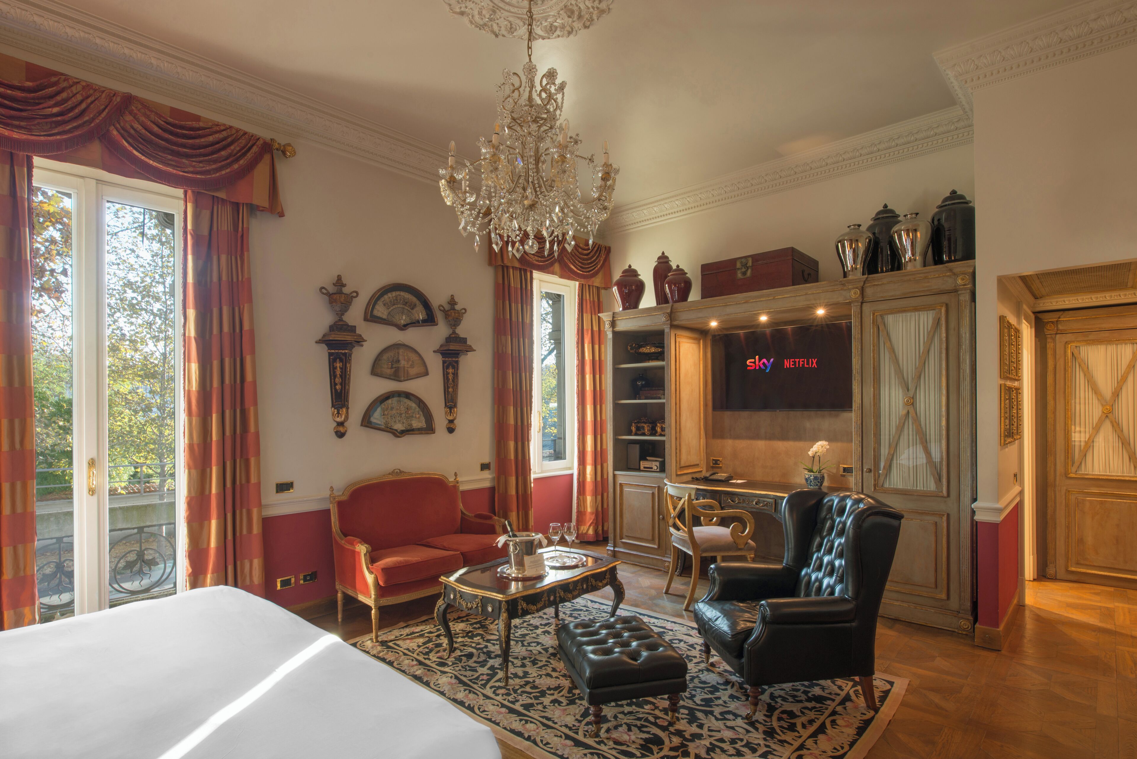 Um quarto com uma cama, um sofá vermelho, uma poltrona preta, um candelabro e um armário com objetos decorativos.