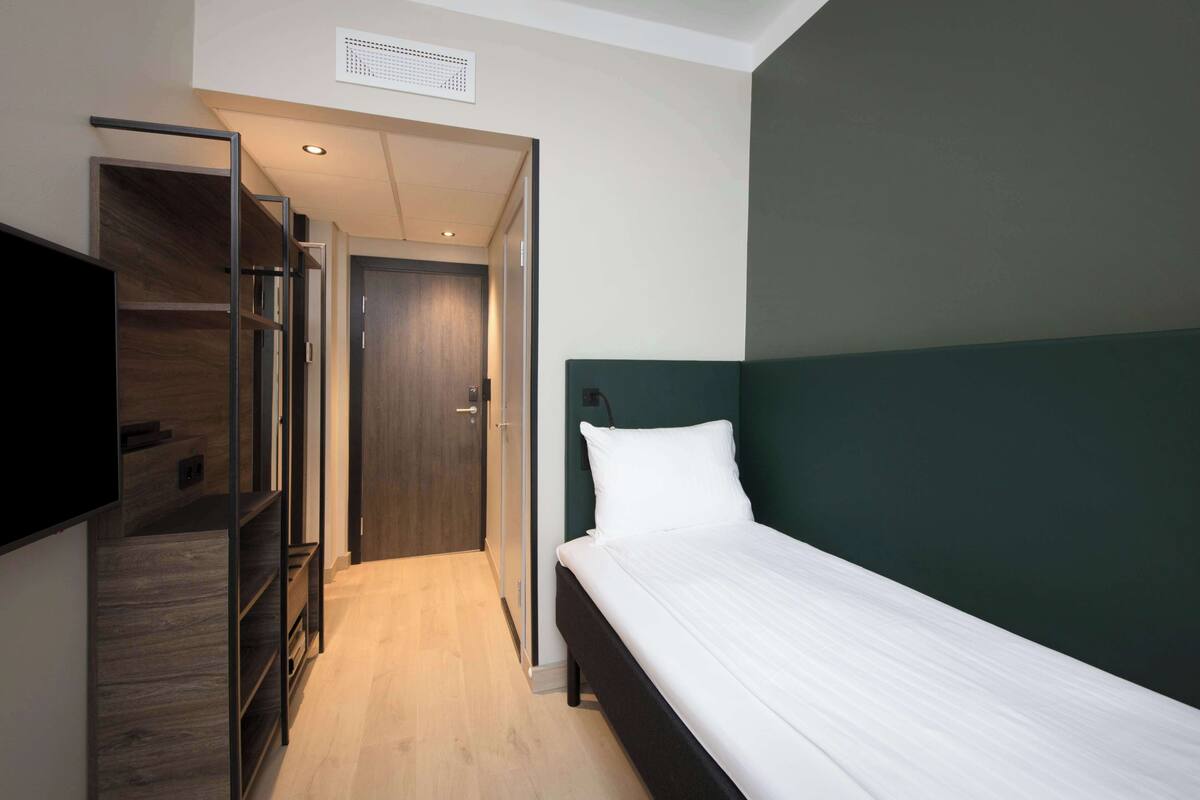Quarto de hotel com cama, televisão, guarda-roupa e porta que dá para outro cômodo.