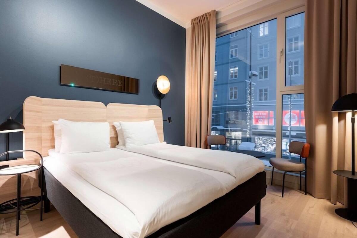 Quarto de hotel com uma cama grande, uma mesinha de cabeceira, uma cadeira, uma janela com vista para prédios e um luminário fixado na parede.