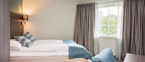 Quarto de hotel com cama, mesa de cabeceira, luminária e janela com cortinas.