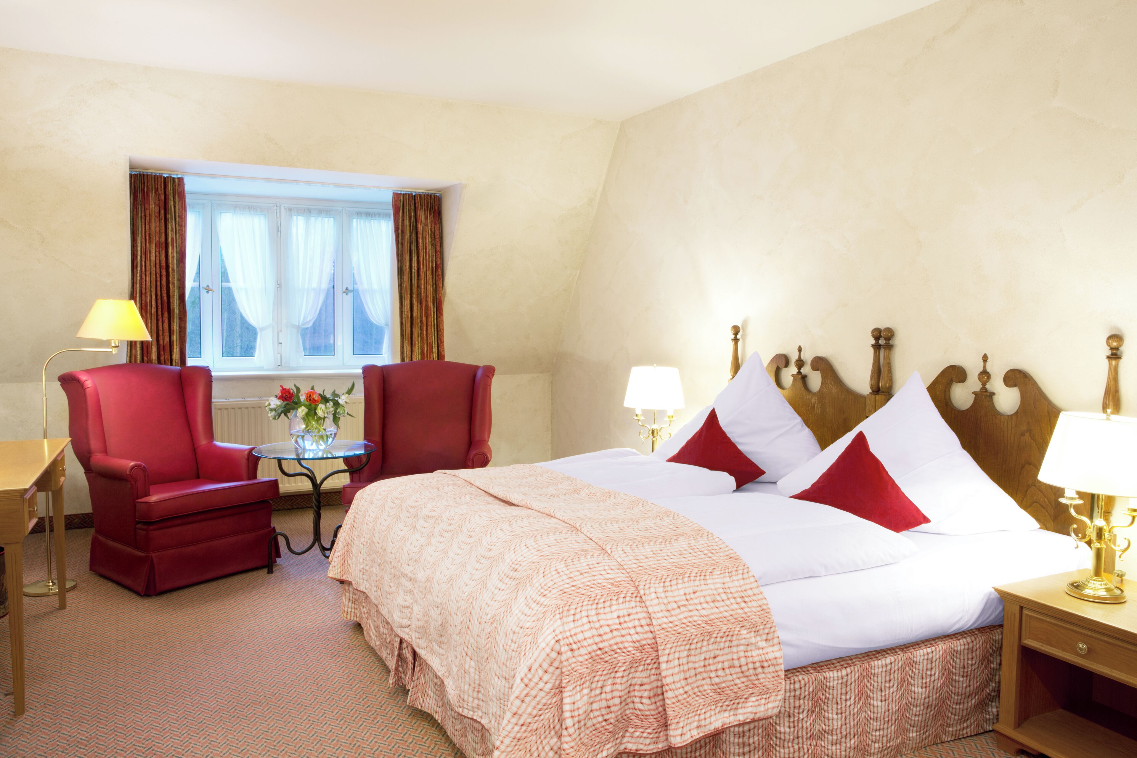 Quarto de hotel com uma cama grande, dois poltronas vermelhas, uma mesa pequena com abajur e uma janela com cortinas.