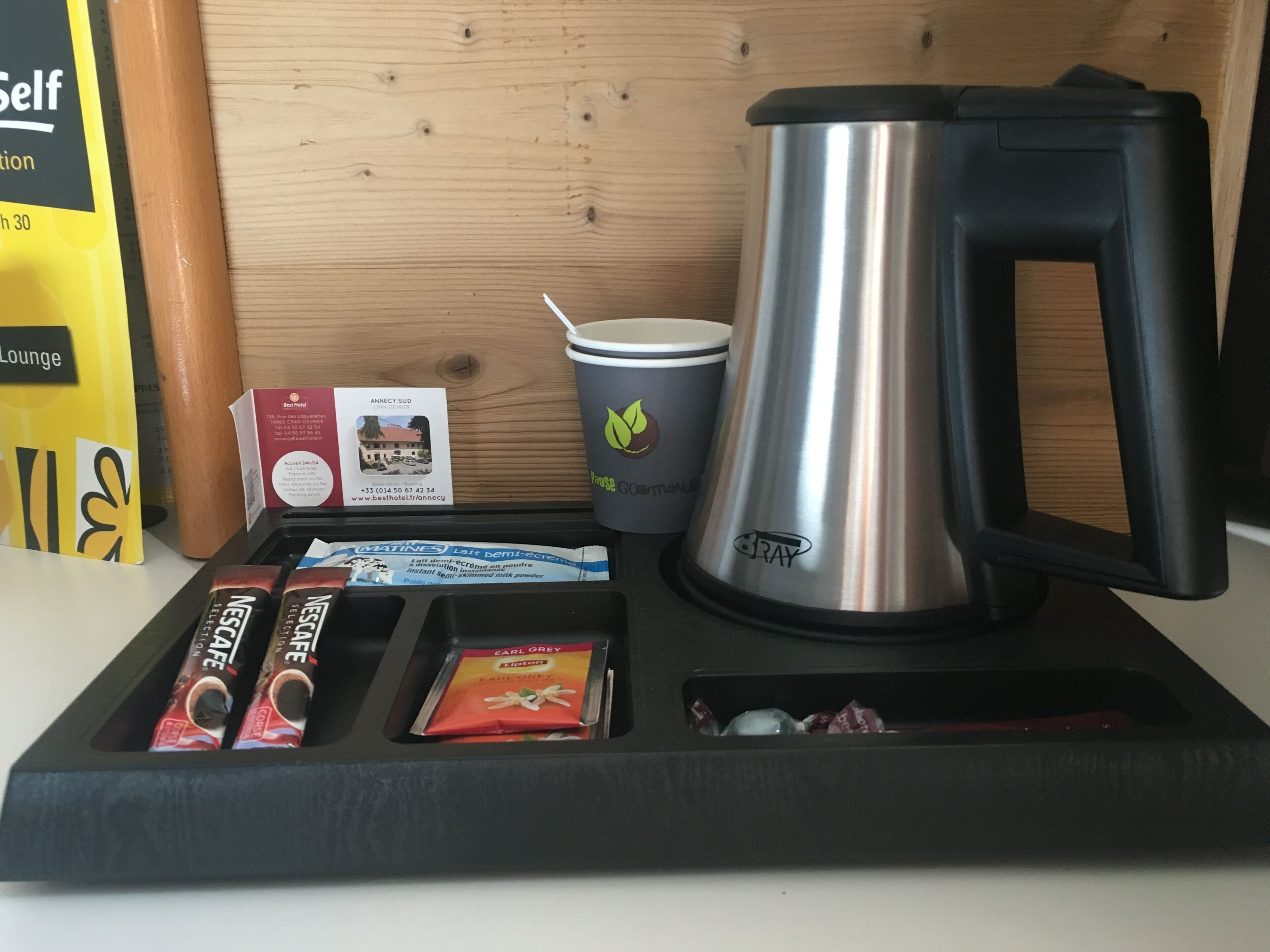 Uma cafeteira, uma chávena, uma embalagem de Nescafe e um folheto sobre uma mesa de madeira.