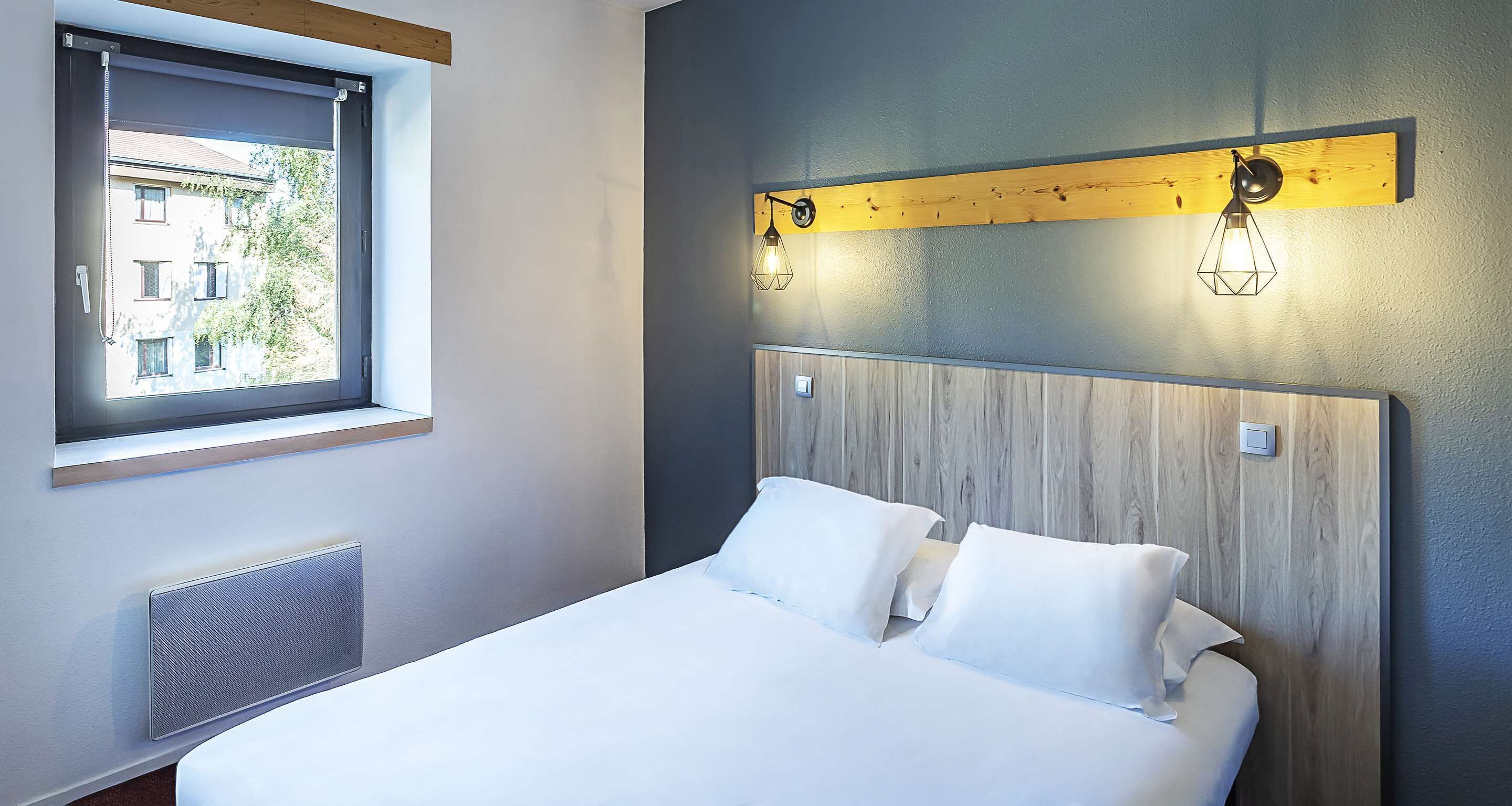 Quarto de hotel com uma cama, uma janela e duas luzes fixadas na parede.
